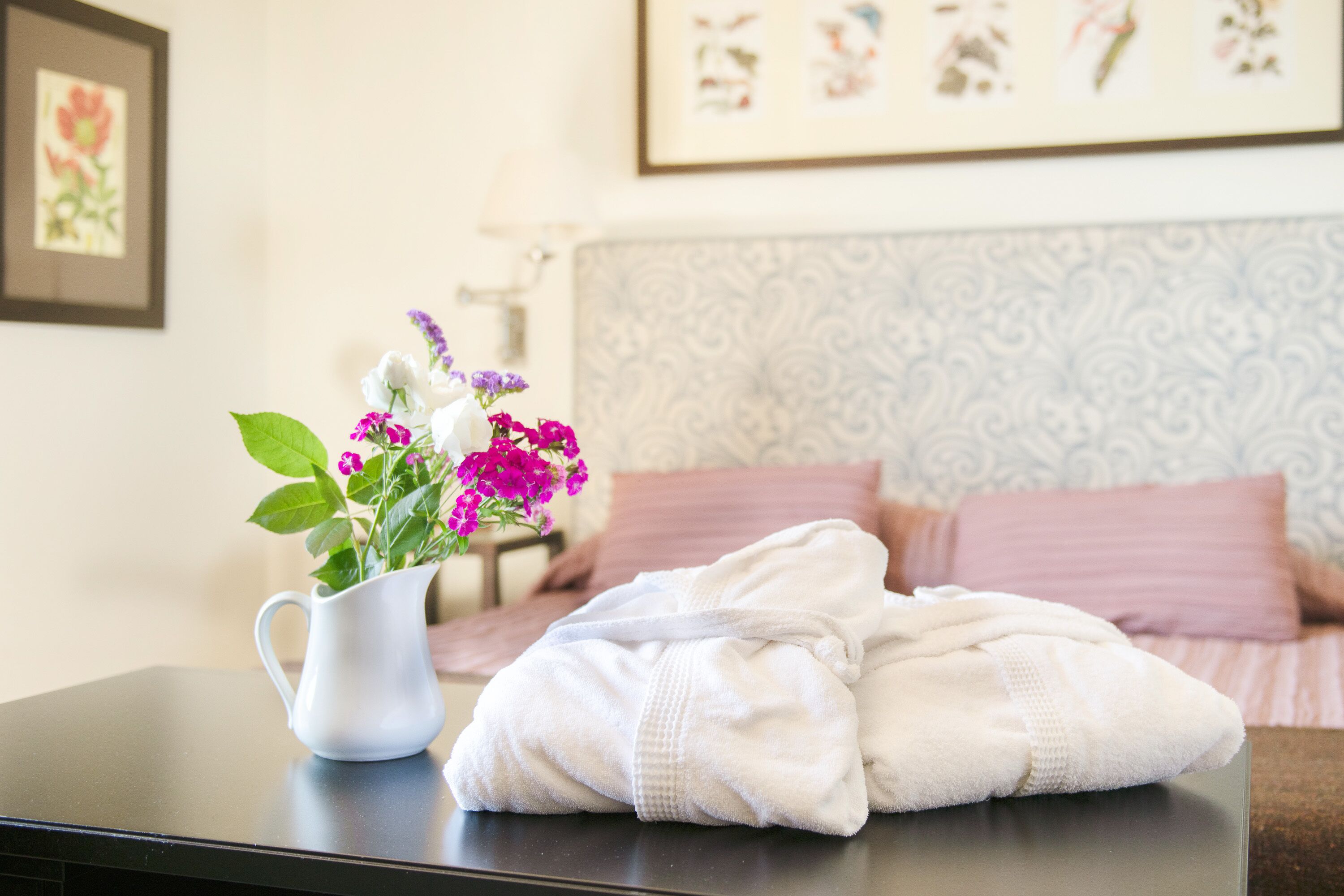 Cama bem arrumada com roupa de cama rosa, roupão branco e um jarro branco com flores sobre uma mesa.