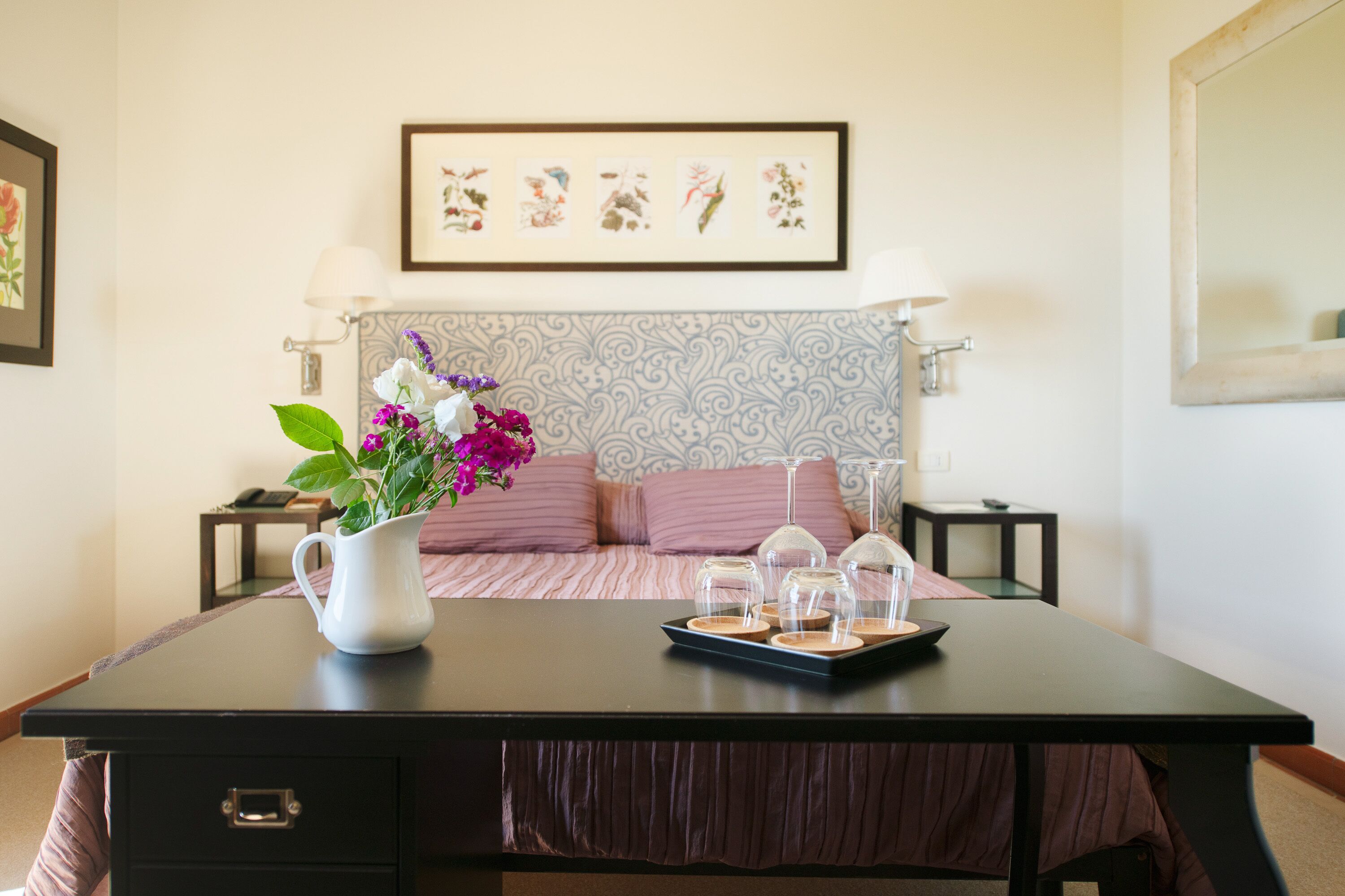 Quarto de hotel com uma cama, mesa de cabeceira com um vaso de flores, luminária e uma pintura floral emoldurada sobre a cama.