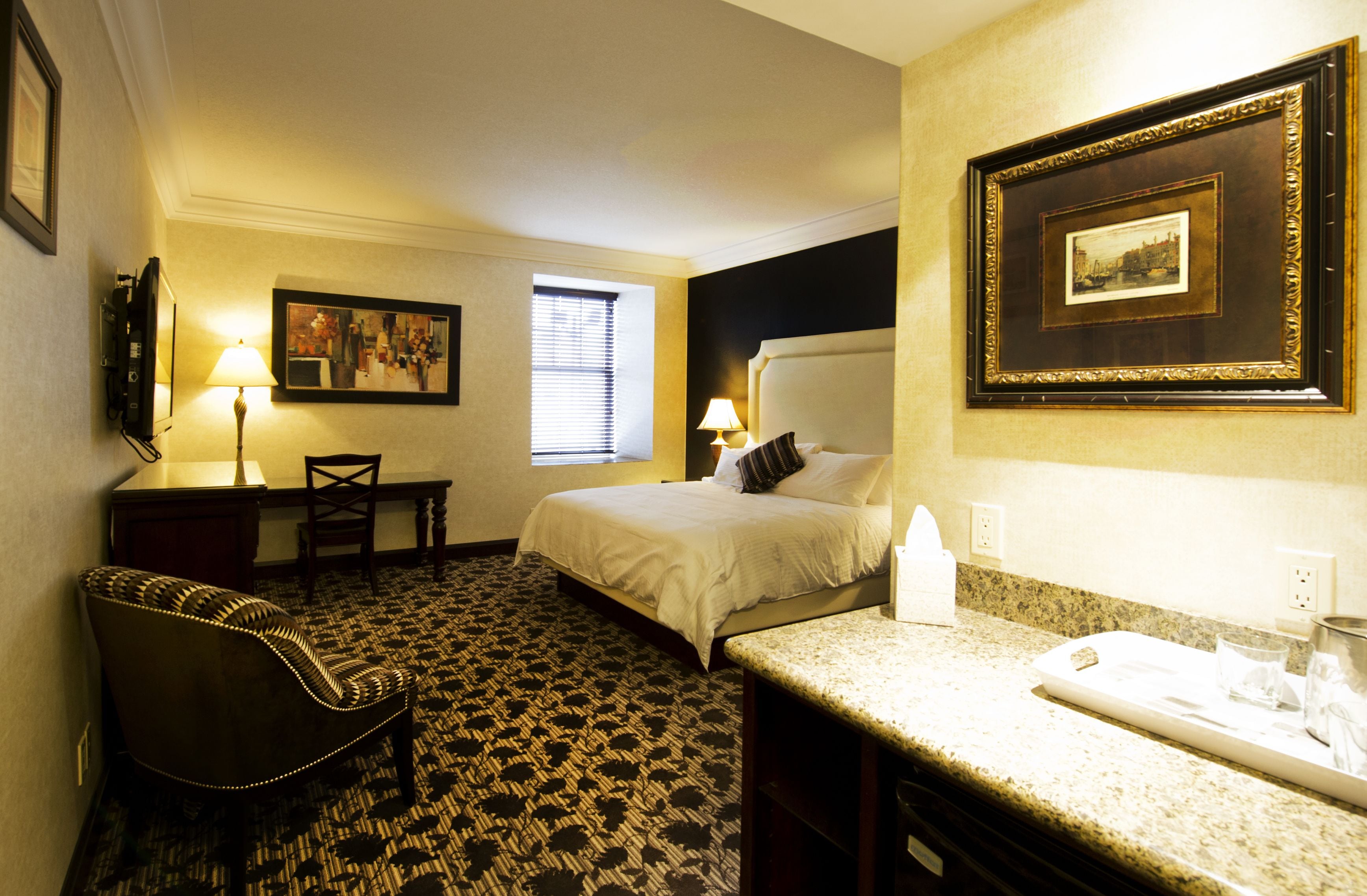Quarto de hotel com cama, escrivaninha, cadeira, televisão e quadro na parede.