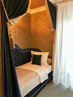 Uma cama com dossel, tendo um painel de cabeceira escuro, um edredom acolchoado e duas almofadas. O quarto apresenta papel de parede estampado e uma grande janela com cortinas brancas.