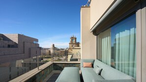 Varanda com sofá e uma mesa pequena, com vista para a paisagem urbana, com edifícios e um campanário.