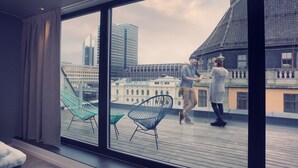 Quarto de hotel moderno com grandes janelas, vista para a paisagem urbana e duas cadeiras verdes na varanda.