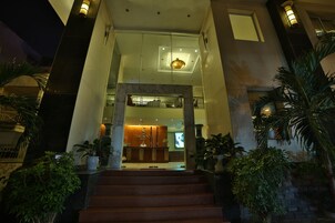 Entrada de hotel com balcão de recepção, escada e plantas em vasos.