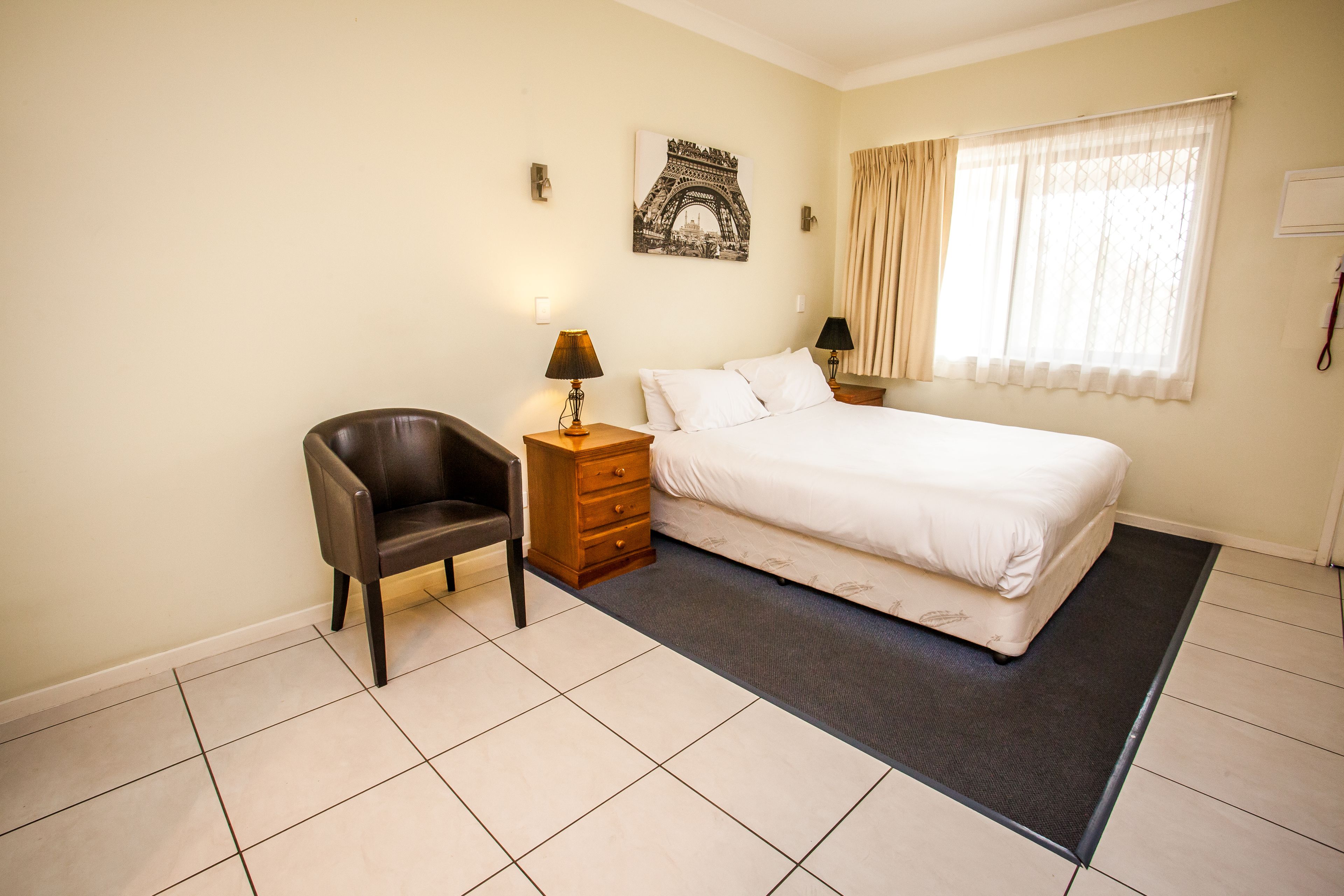 Um quarto com cama, uma cadeira, mesa de cabeceira, abajur, janela com cortinas e um quadro na parede da Torre Eiffel.