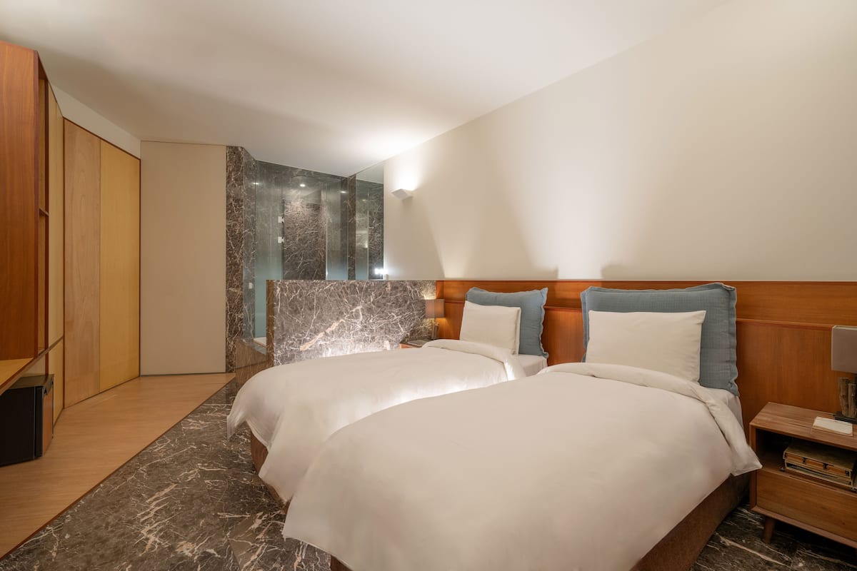 Quarto de hotel com duas camas, guarda-roupa de madeira, parede de mármore e mesa de cabeceira com abajur.