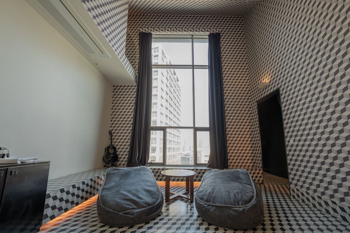 Um quarto com piso xadrez, duas poltronas de grão de espuma, uma pequena mesa e uma janela com vista para edifícios altos.