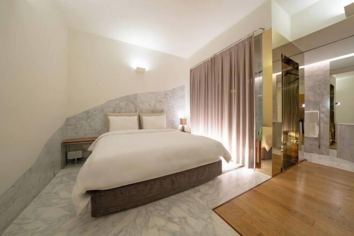 Quarto de hotel moderno com parede em mármore, uma cama grande, piso de madeira e divisória de vidro.