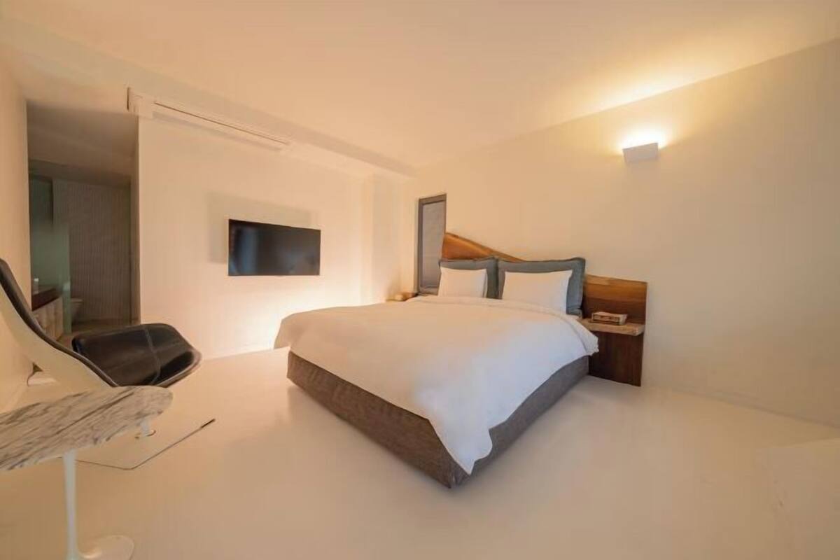 Quarto de hotel moderno com uma cama grande, uma mesa de cabeceira, uma cadeira e uma televisão de tela plana na parede.