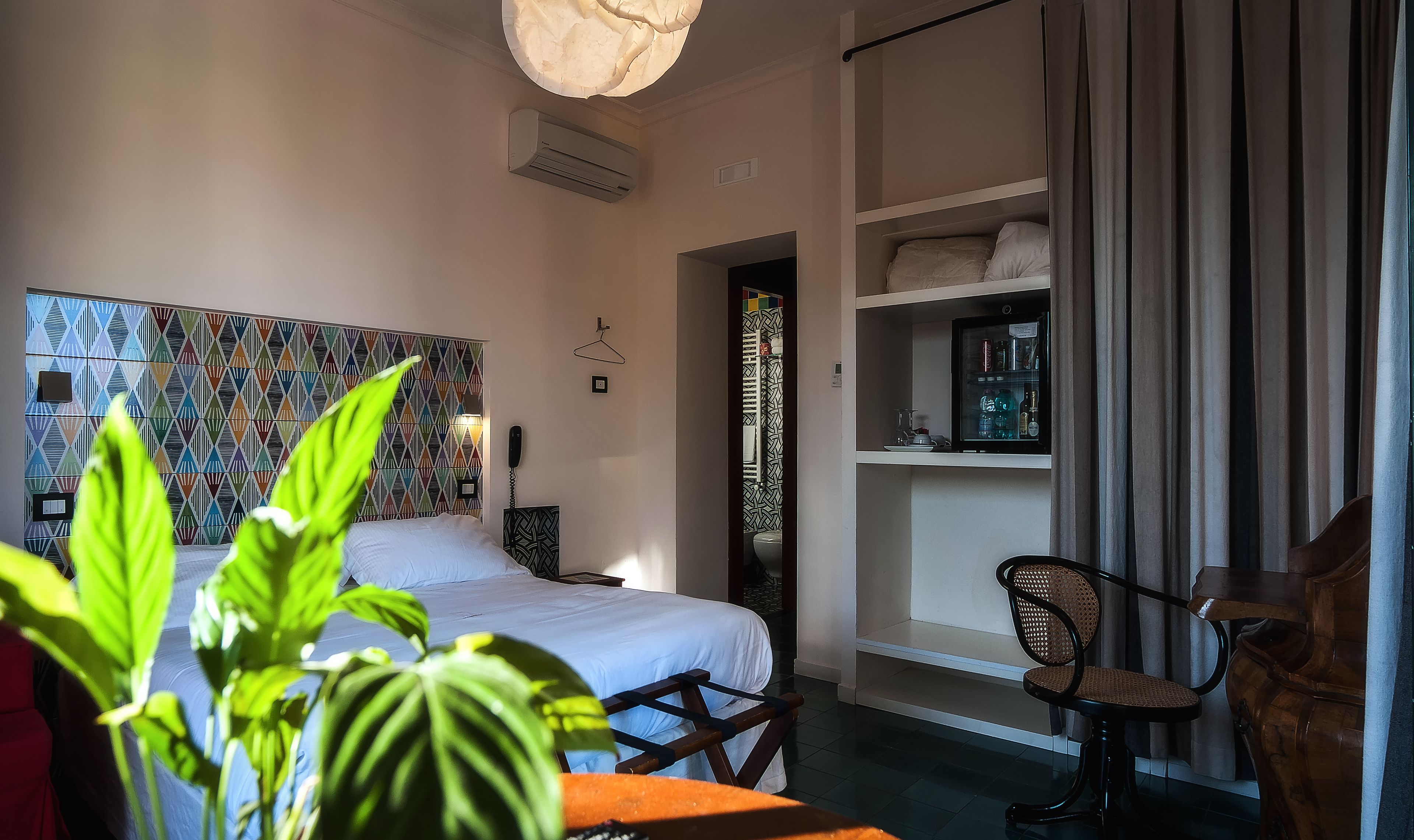 Quarto de hotel com cama, uma cadeira, prateleira com garrafas e parede estampada.