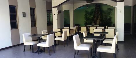 Uma área de refeições com mesas e cadeiras, uma parede verde com plantas e piso de azulejos.
