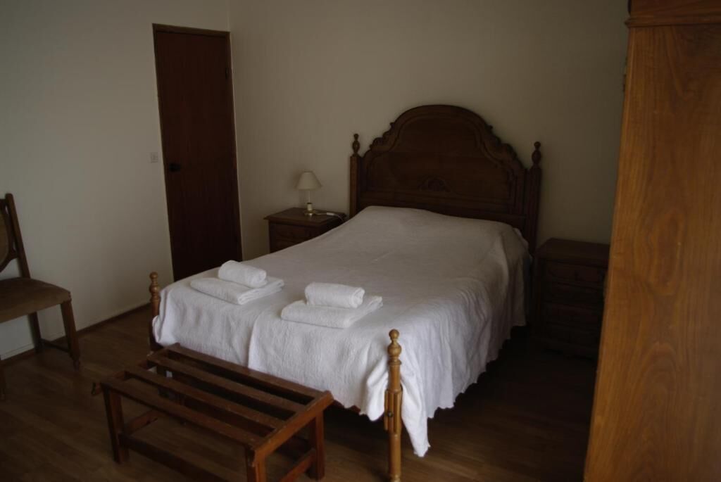 Quarto com cama de madeira, mesa de cabeceira com abajur, um banco de madeira e uma porta.