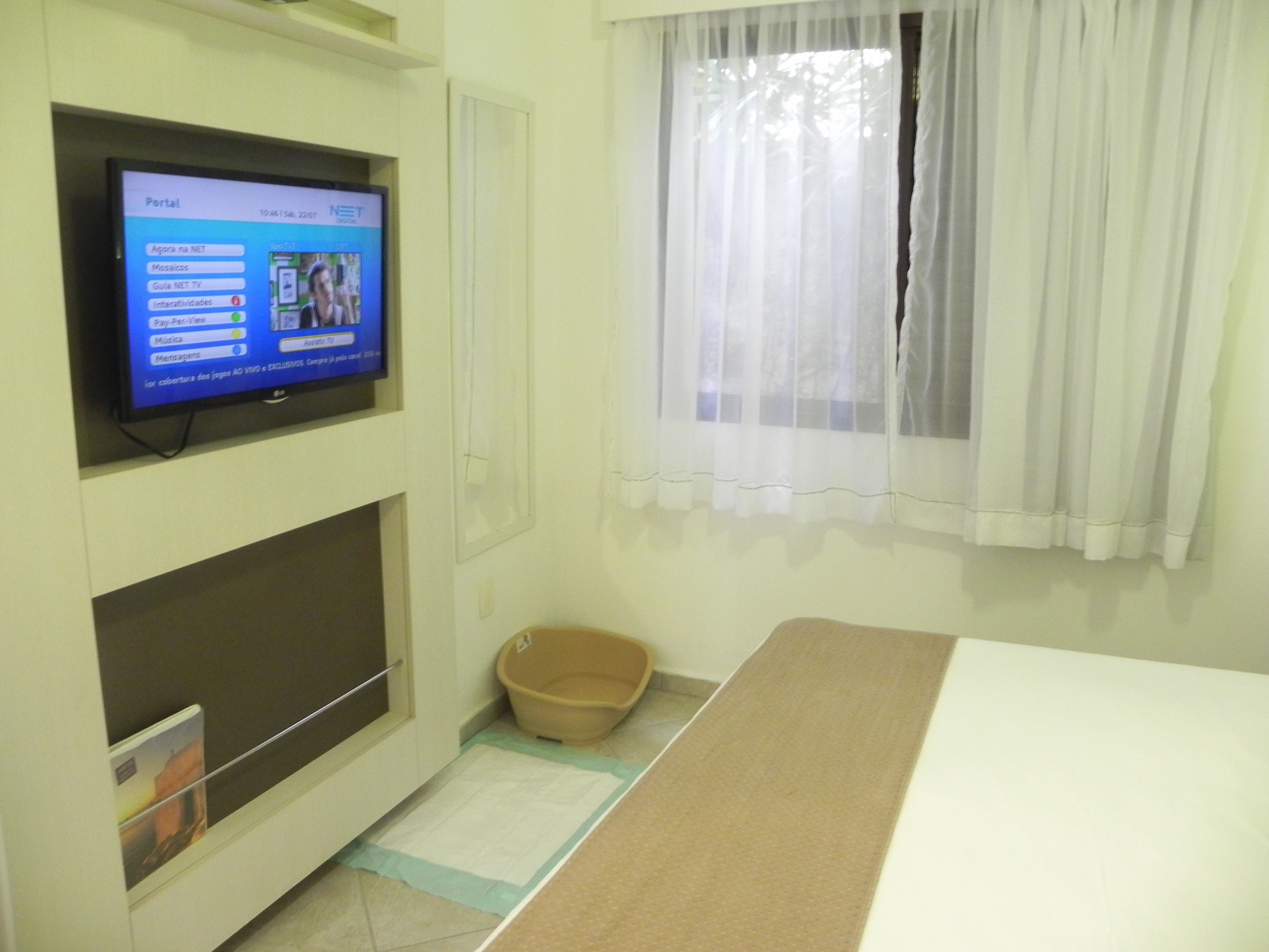 Quarto de hotel com televisão na parede exibindo uma interface de portal, uma janela pequena com cortinas leves, uma banheira bege e uma cama com colcha marrom.