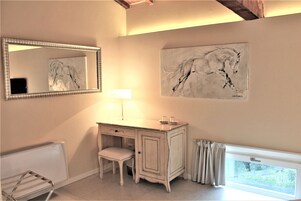 Quarto com escrivaninha branca, cadeira e um espelho grande. Há um quadro de um cavalo na parede e uma janela com cortinas.