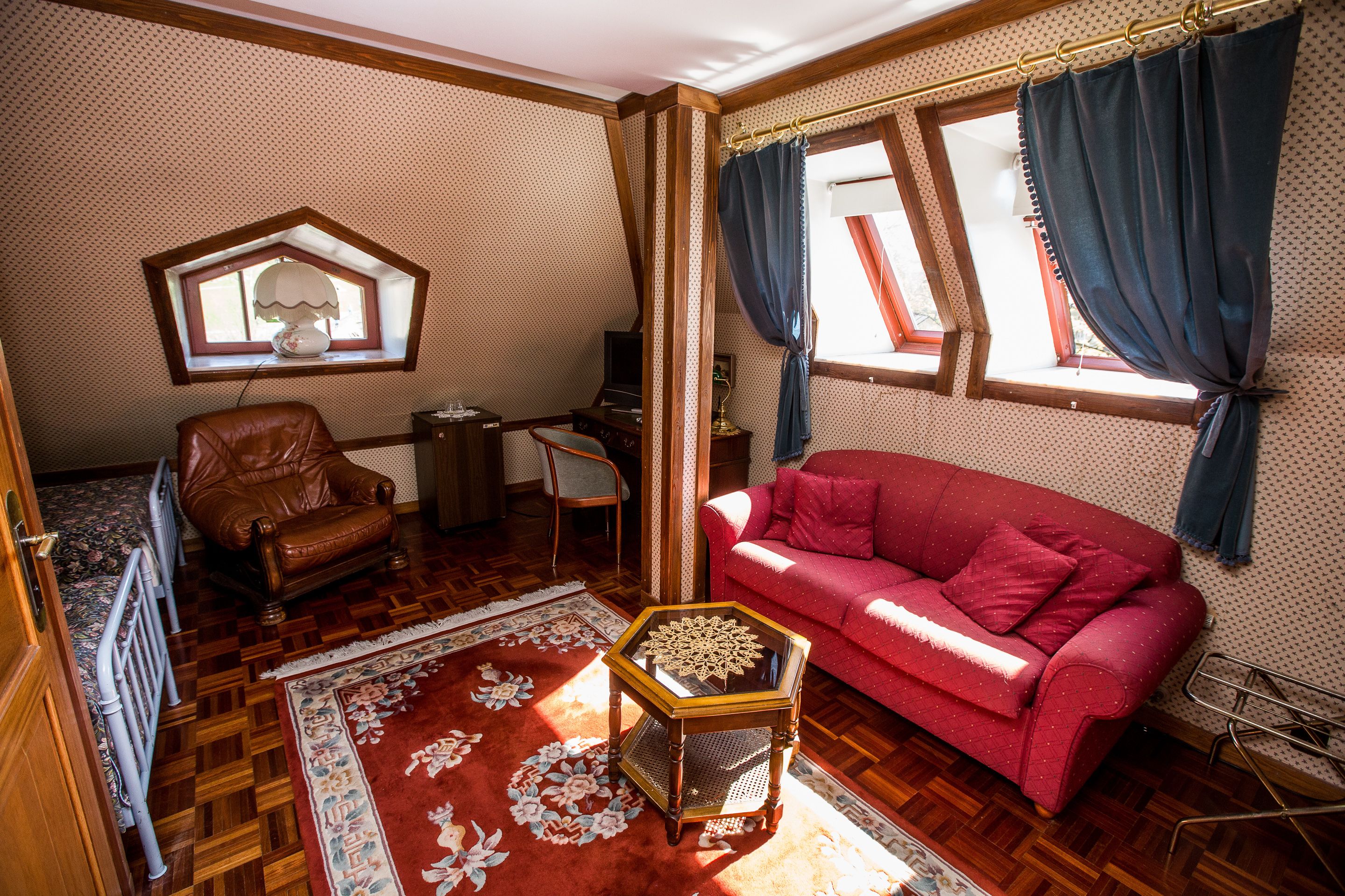 Quarto com sofá vermelho, poltrona de couro, mesa de centro de madeira, tapete estampado e janela com cortinas.