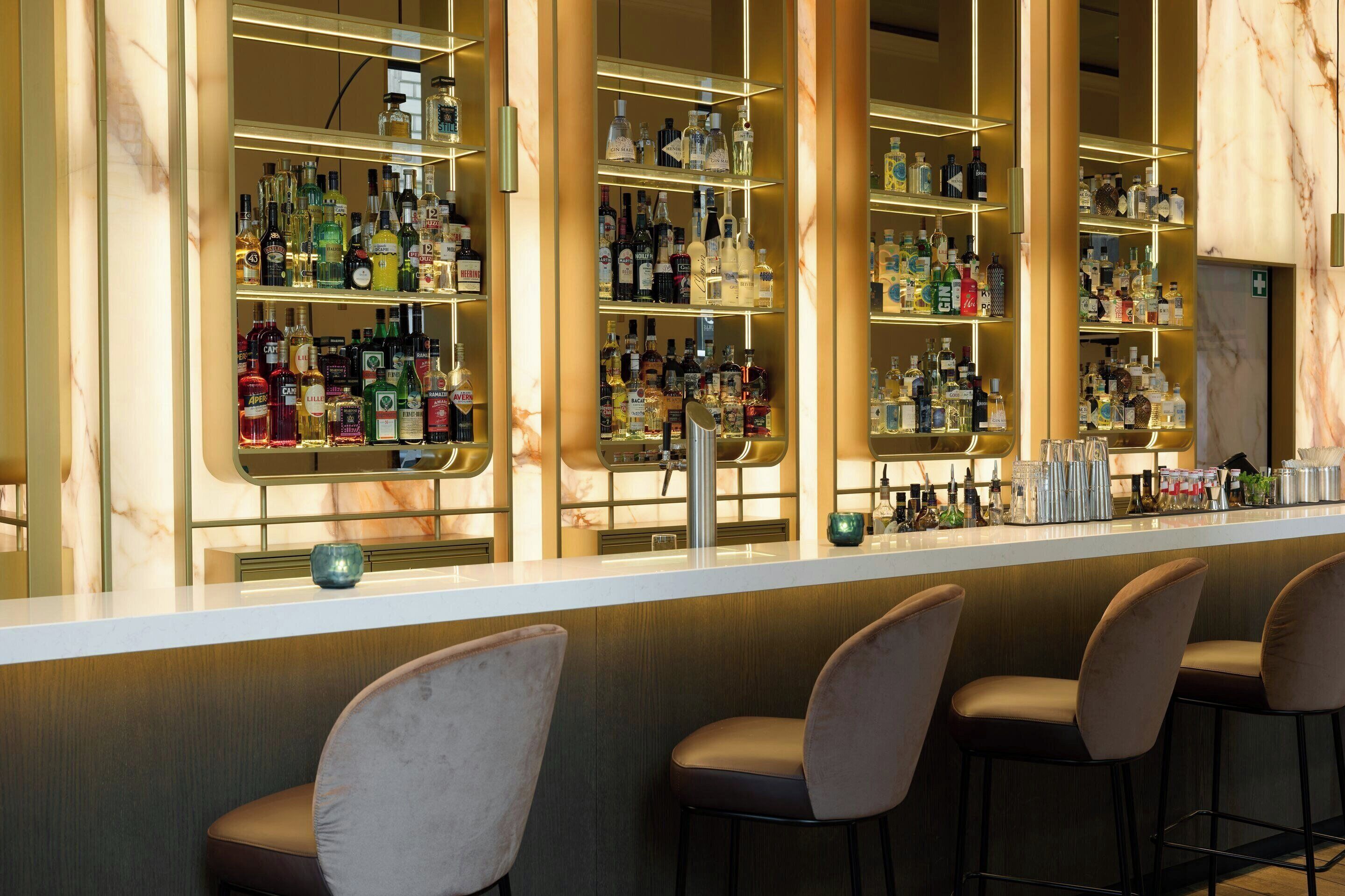 Uma área de bar com várias prateleiras de garrafas de bebidas, um balcão e banquetas.