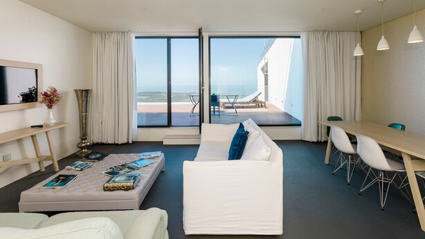 Quarto de hotel moderno com uma grande porta de vidro deslizante que dá para uma varanda, um sofá branco, uma mesinha e uma TV fixada na parede.