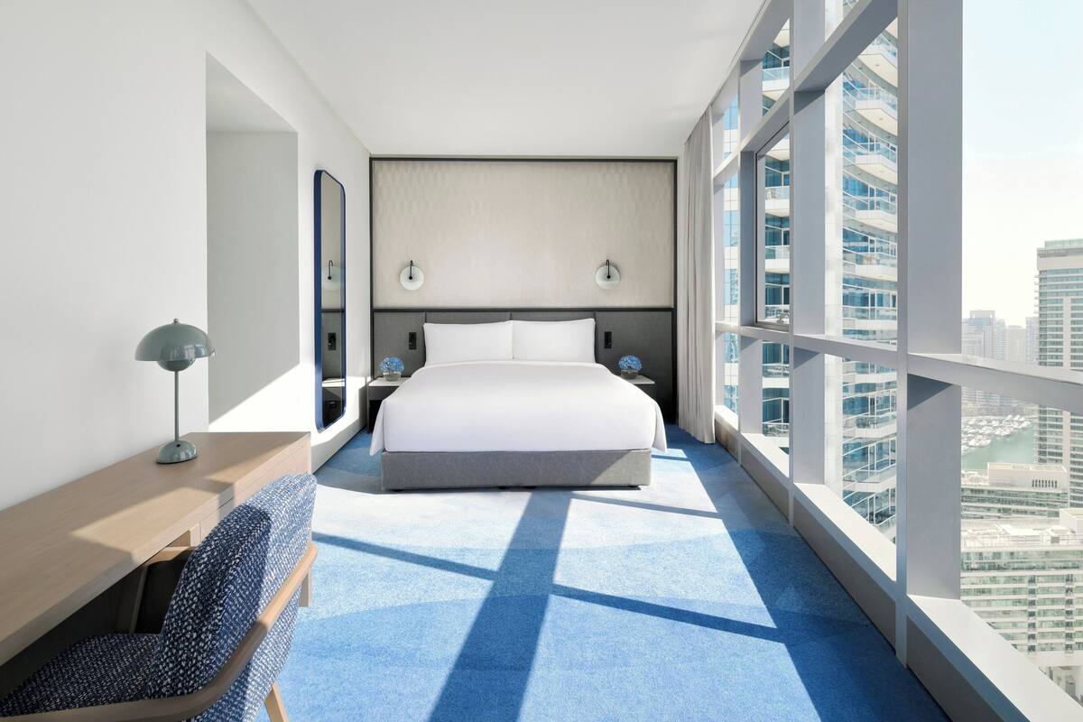 Quarto de hotel moderno com uma cama grande, uma escrivaninha, uma cadeira, uma mesa de cabeceira e vista para a cidade através de amplas janelas.