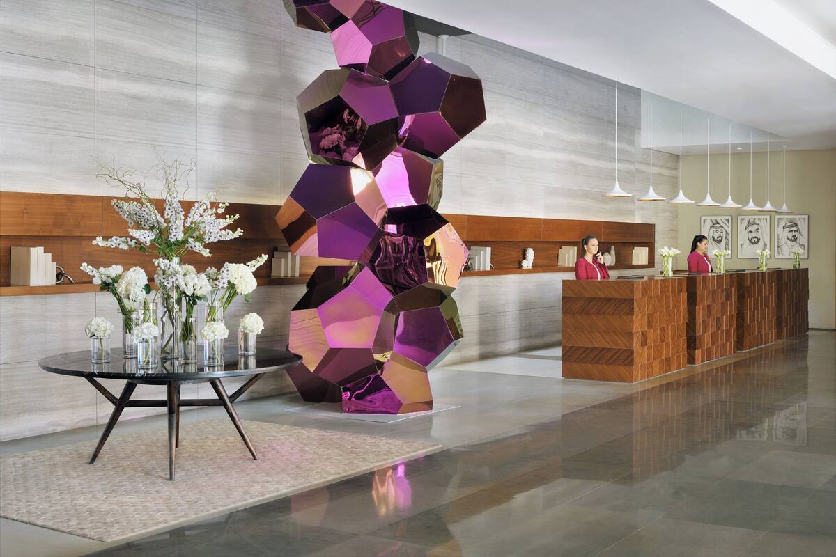 Uma área de recepção com um balcão moderno, uma escultura geométrica decorativa e um vaso com flores brancas.