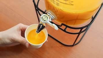 Uma mão despejando líquido laranja de um dispensador de metal em uma xícara branca.