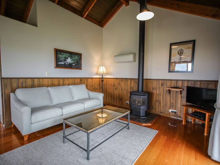 Uma sala de estar aconchegante com um sofá branco, uma mesa de centro de vidro, uma lareira a lenha, uma televisão sobre um suporte de madeira e quadros nas paredes.