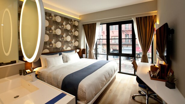 Quarto de hotel com uma cama grande, uma escrivaninha, uma cadeira, uma luminária de mesa, um espelho e uma janela com cortinas.