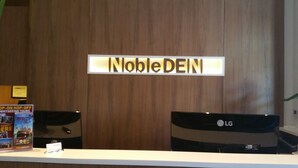 Uma mesa de recepção com uma placa indicando "Noble DEN" e dois monitores LG.