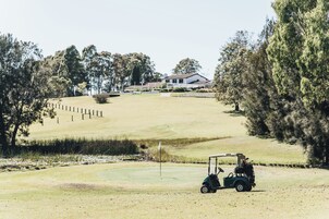 Um campo de golfe com um carrinho de golfe, árvores e uma casa ao fundo.