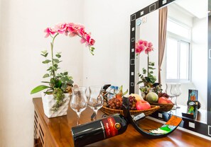 Uma mesa de madeira com uma garrafa de vinho do Porto, um vaso branco com flores cor-de-rosa, uma cesta de frutas e um espelho refletindo o quarto.