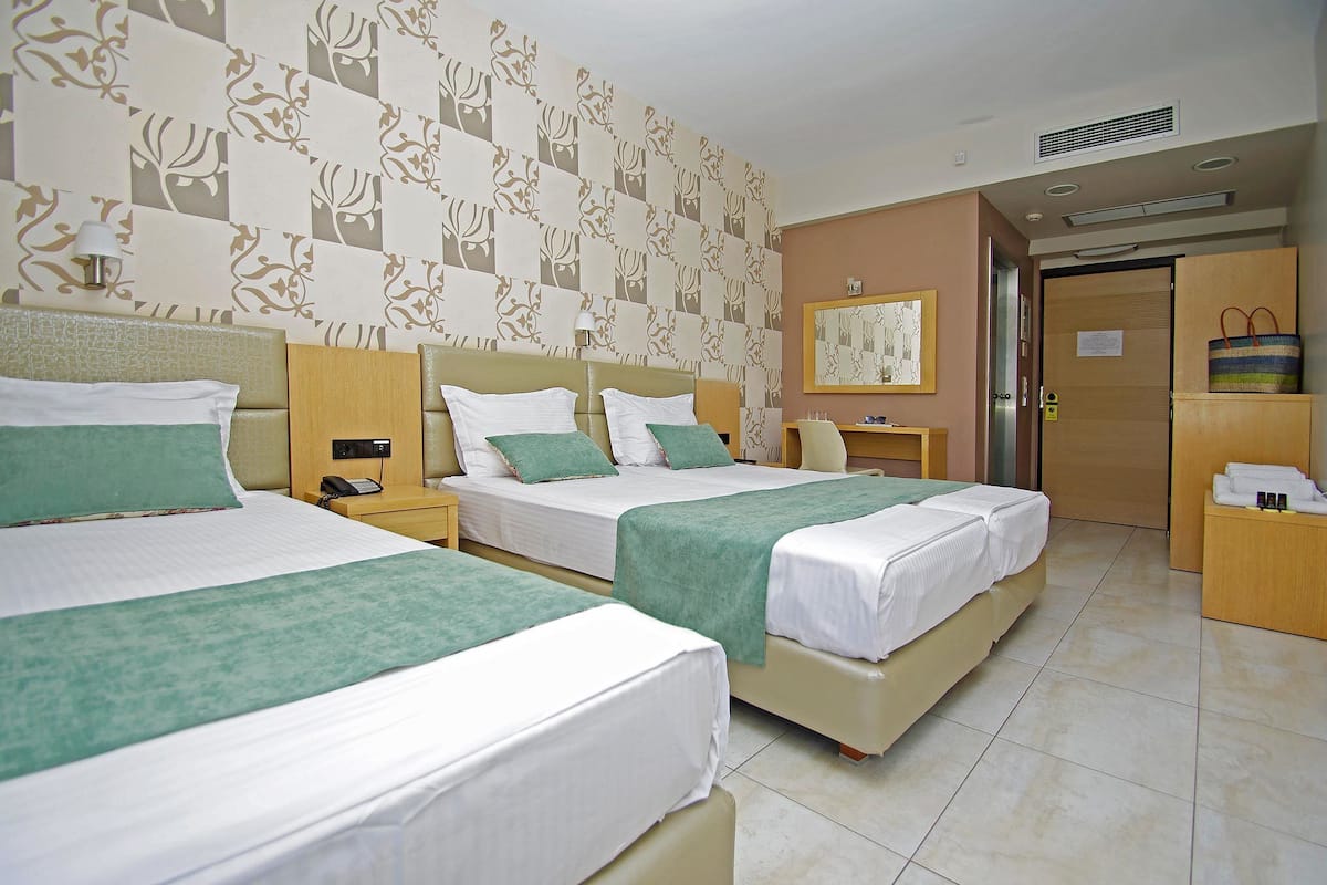 Quarto de hotel com duas camas, um guarda-roupa de madeira, um espelho e uma porta que dá para outro cômodo.