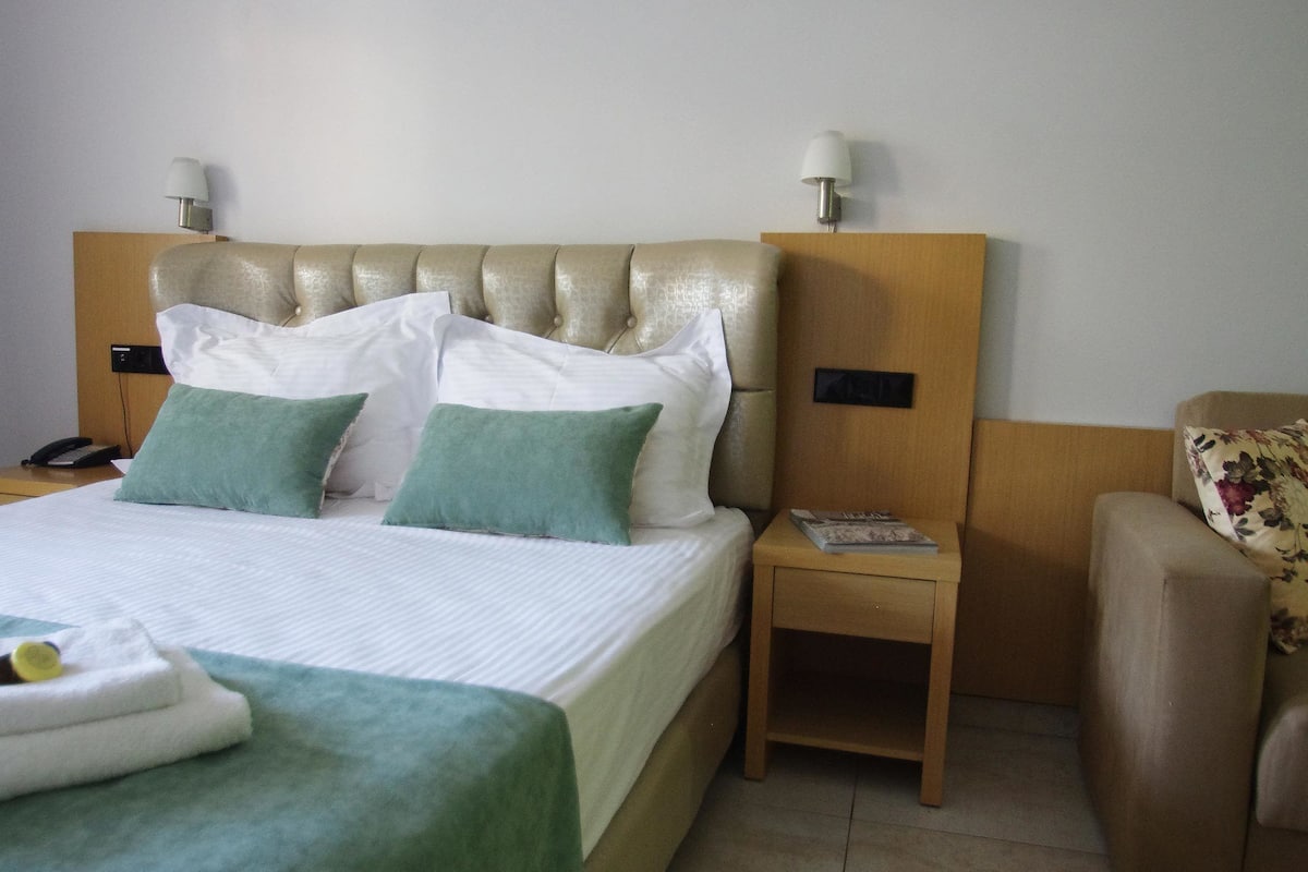 Quarto de hotel com uma cama, duas mesas de cabeceira, um criado-mudo, uma cadeira e um abajur.