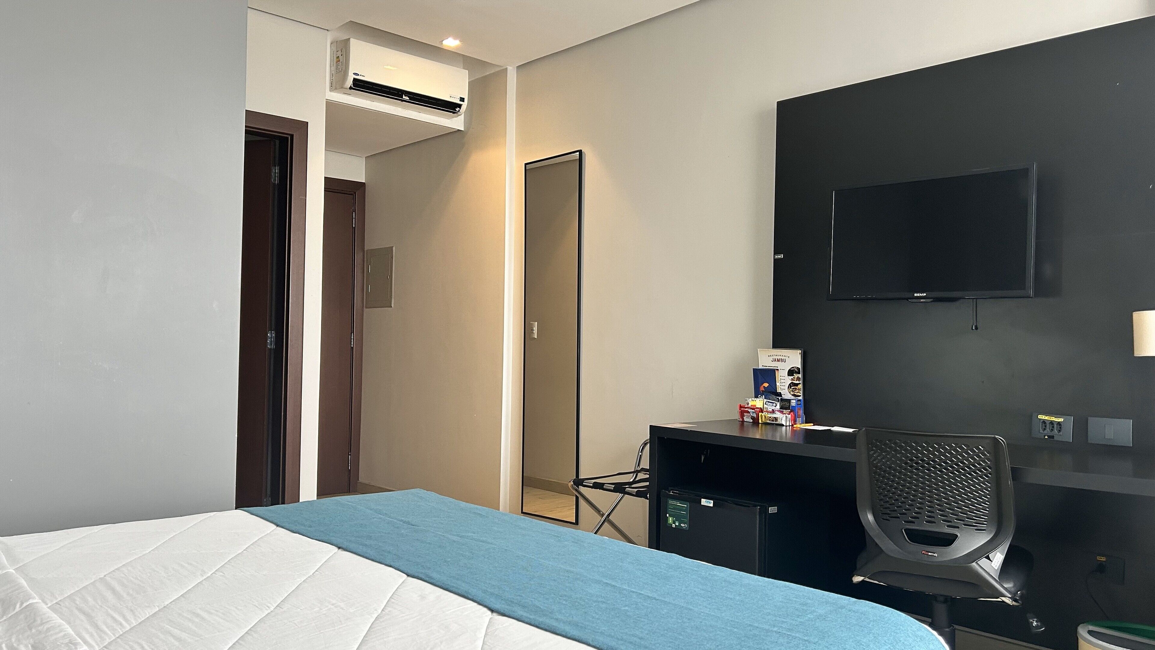 Quarto de hotel com cama, escrivaninha com cadeira, televisão de tela plana, ar condicionado e espelho.