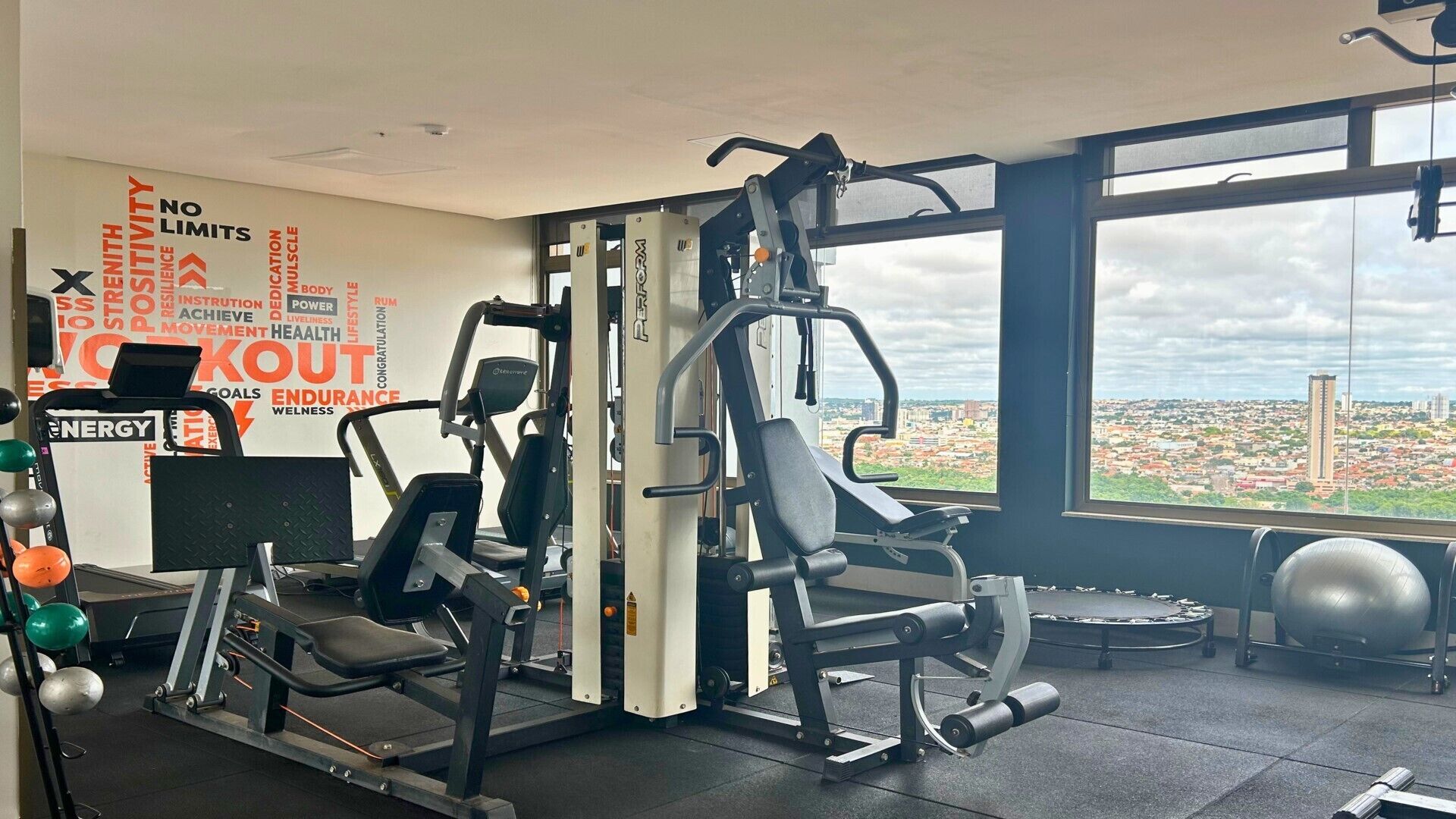 Uma academia com diversos equipamentos de exercício, uma parede com mensagens motivacionais e vista para o horizonte urbano através de amplas janelas.