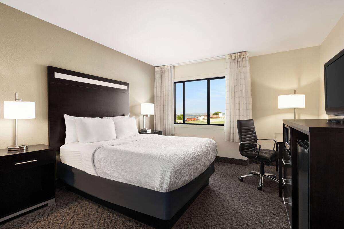 Quarto de hotel com uma cama grande, uma mesa com televisão, uma cadeira e uma janela com vista.