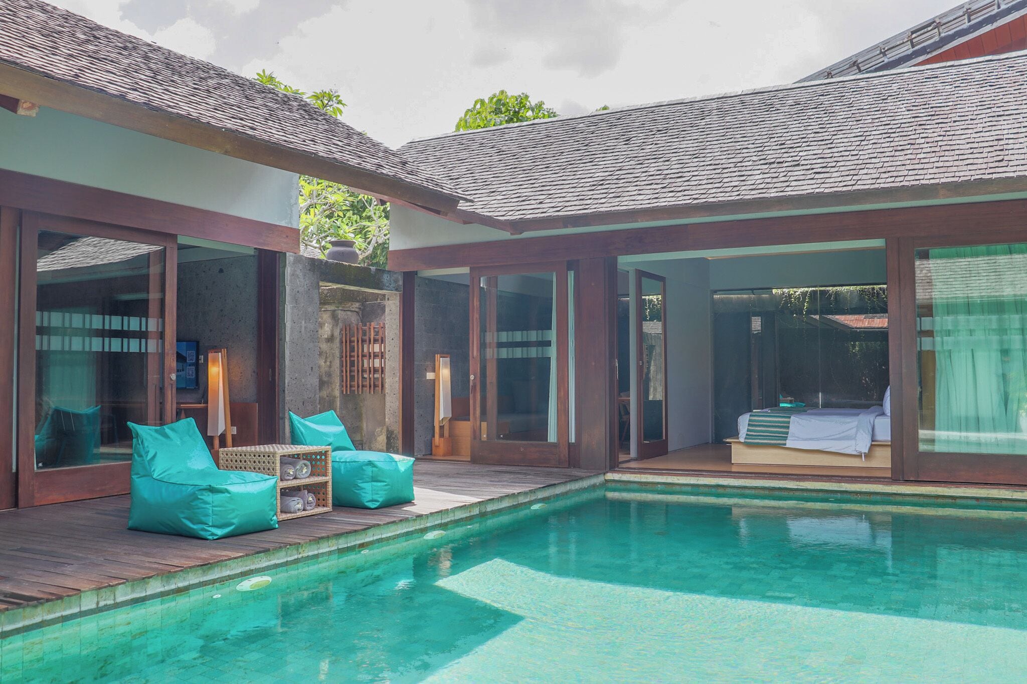 Uma área moderna à beira da piscina, com deck de madeira, sala envidraçada e cama com colcha verde.