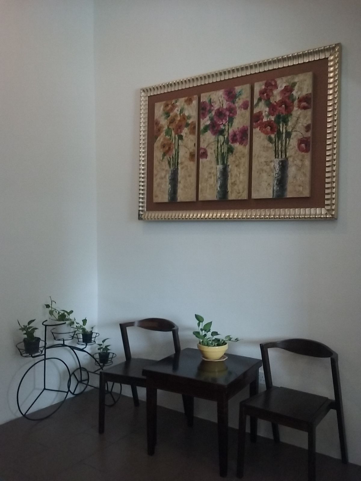 Uma área de refeições com mesa e cadeiras, um quadro de flores pendurado na parede e um suporte de plantas com vasos.