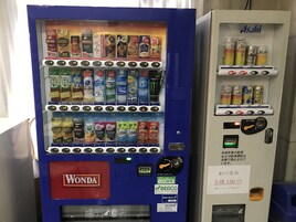 Máquina automática com diversas bebidas e petiscos, incluindo latas de Wonda e Asahi, e um dispensador de dinheiro.