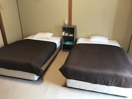 Duas camas de solteiro com lençóis brancos e cabeceiras escuras, uma pequena mesa de cabeceira com abajur e uma prateleira na parede com produtos de higiene pessoal.