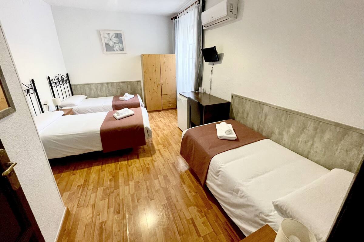 Quarto de hotel com duas camas, um guarda-roupa de madeira, uma escrivaninha com computador e uma televisão.