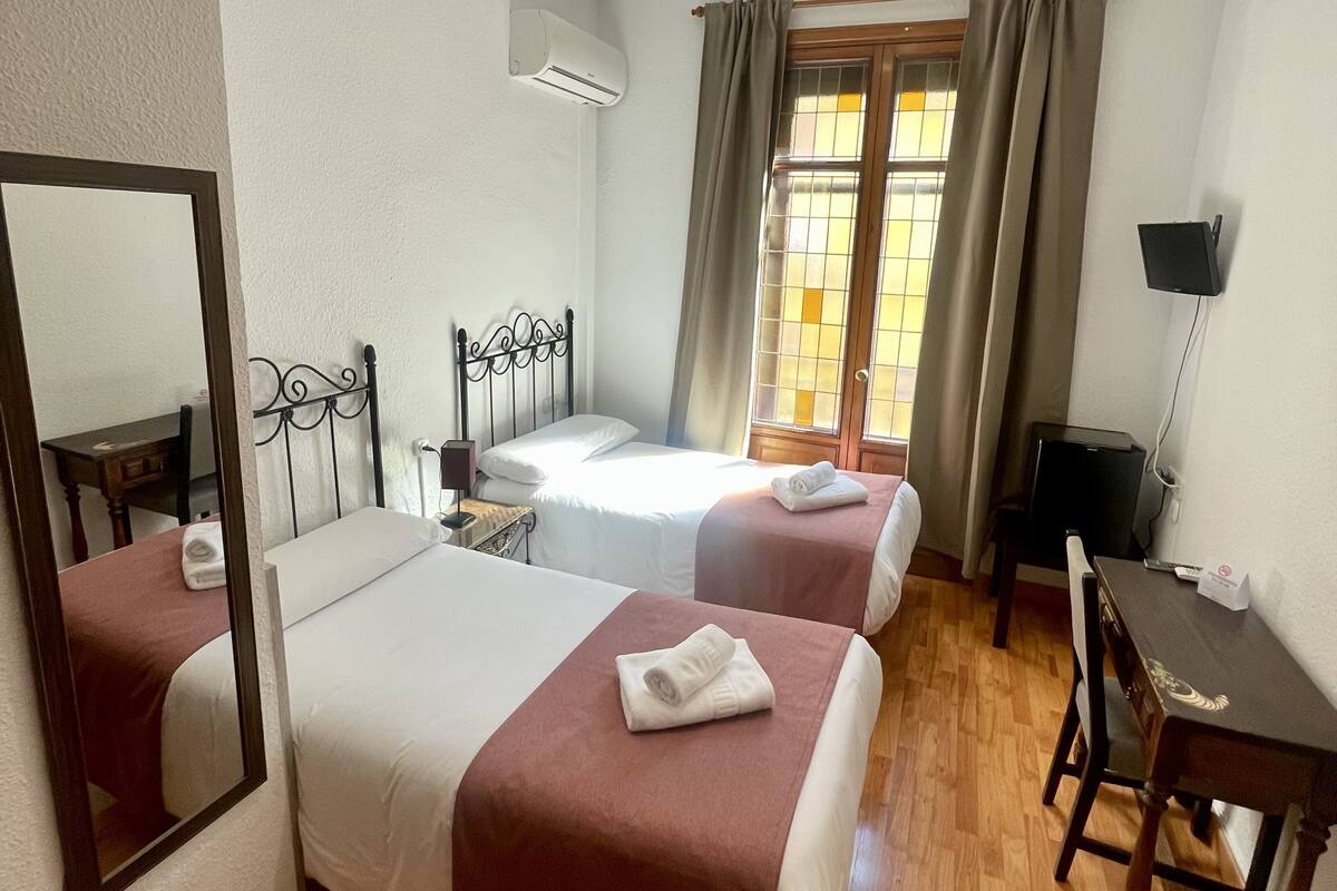 Quarto de hotel com duas camas, uma escrivaninha, uma cadeira, uma televisão e uma janela com cortinas.