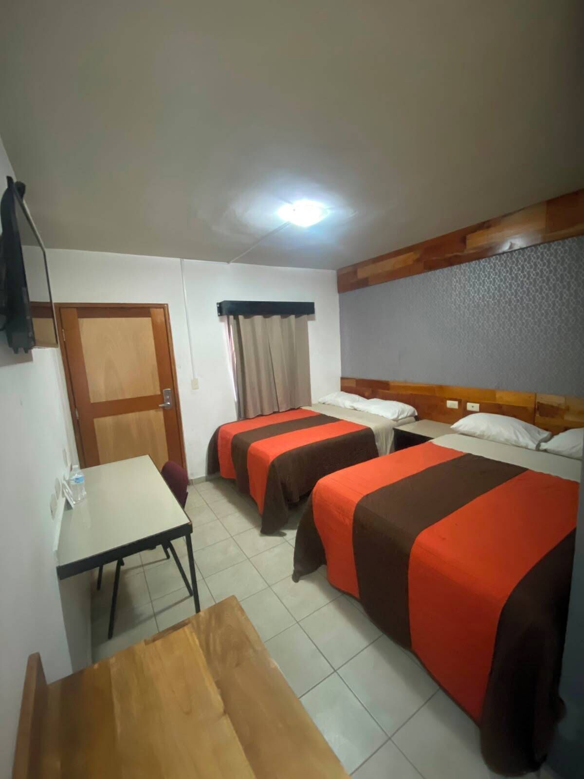 Quarto de hotel com duas camas, uma mesinha, uma cadeira, um banco de madeira, uma escrivaninha e uma porta.