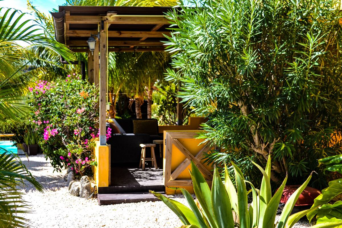 Uma área de estar coberta ao ar livre, com pérgola de madeira, rodeada por vegetação exuberante e plantas floridas.