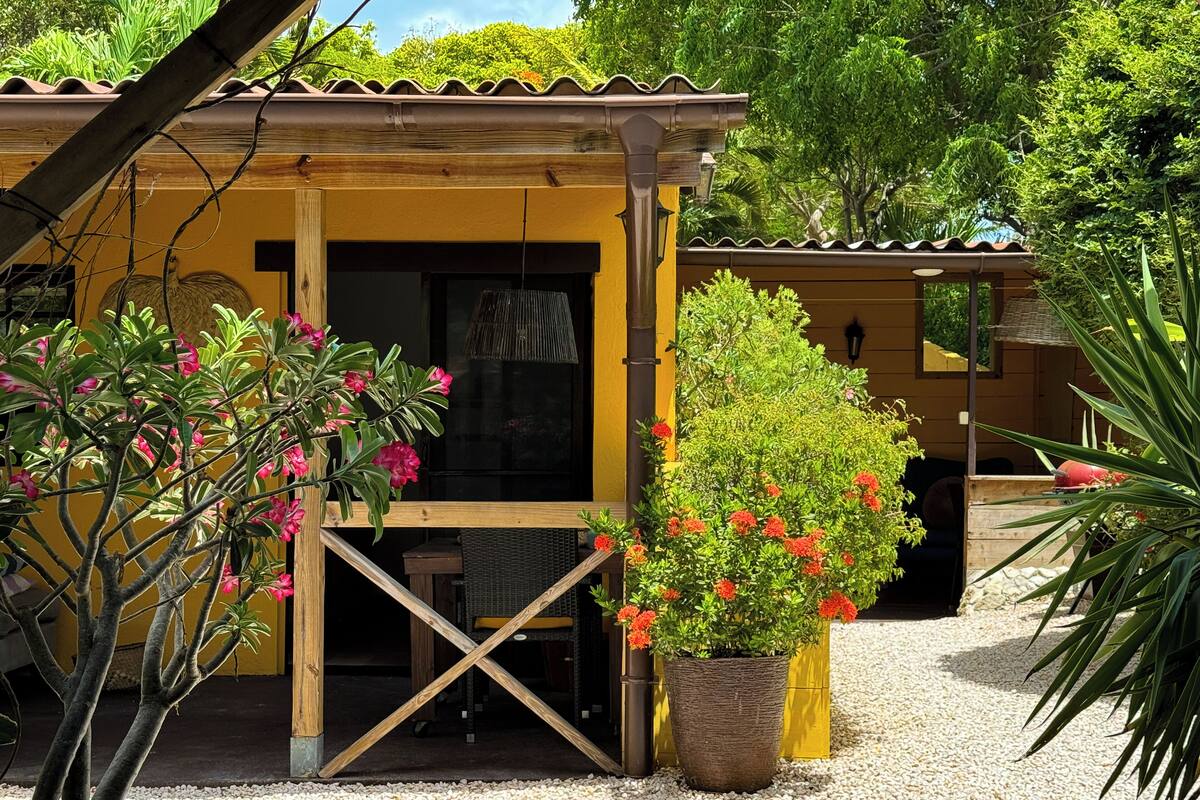 Um edifício amarelo com varanda de madeira e um vaso com flores vermelhas.