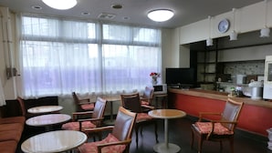 Quarto de hotel com área de refeições, mesas e cadeiras redondas, cozinha equipada com micro-ondas e um relógio, e amplas janelas com cortinas leves.