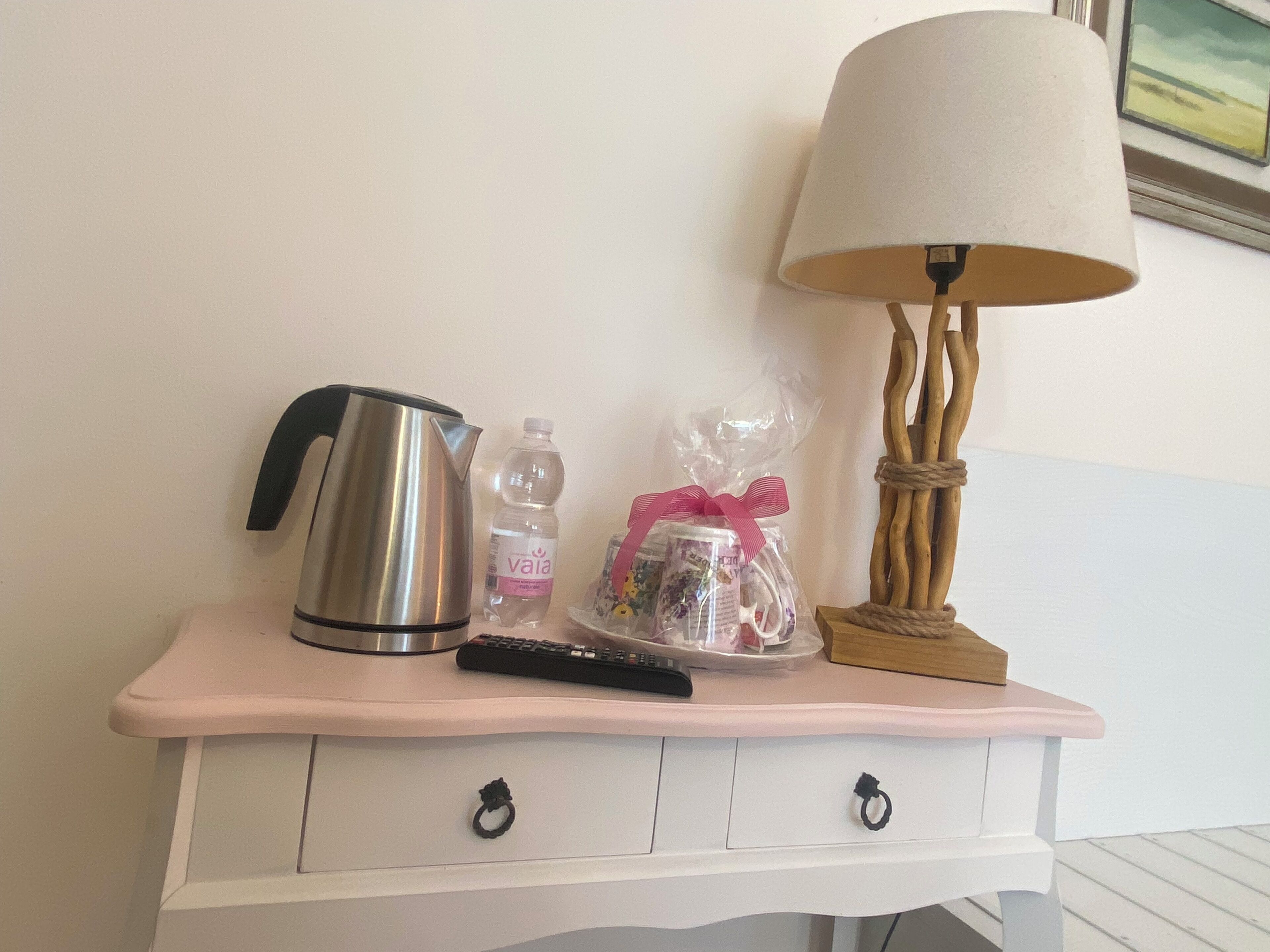 Uma mesa de cabeceira branca com uma lâmpada, uma chaleira, um controle remoto e um pequeno saco plástico com um laço rosa.