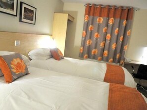 Quarto de hotel com duas camas, mesa de cabeceira, abajur e janela com cortinas florais.