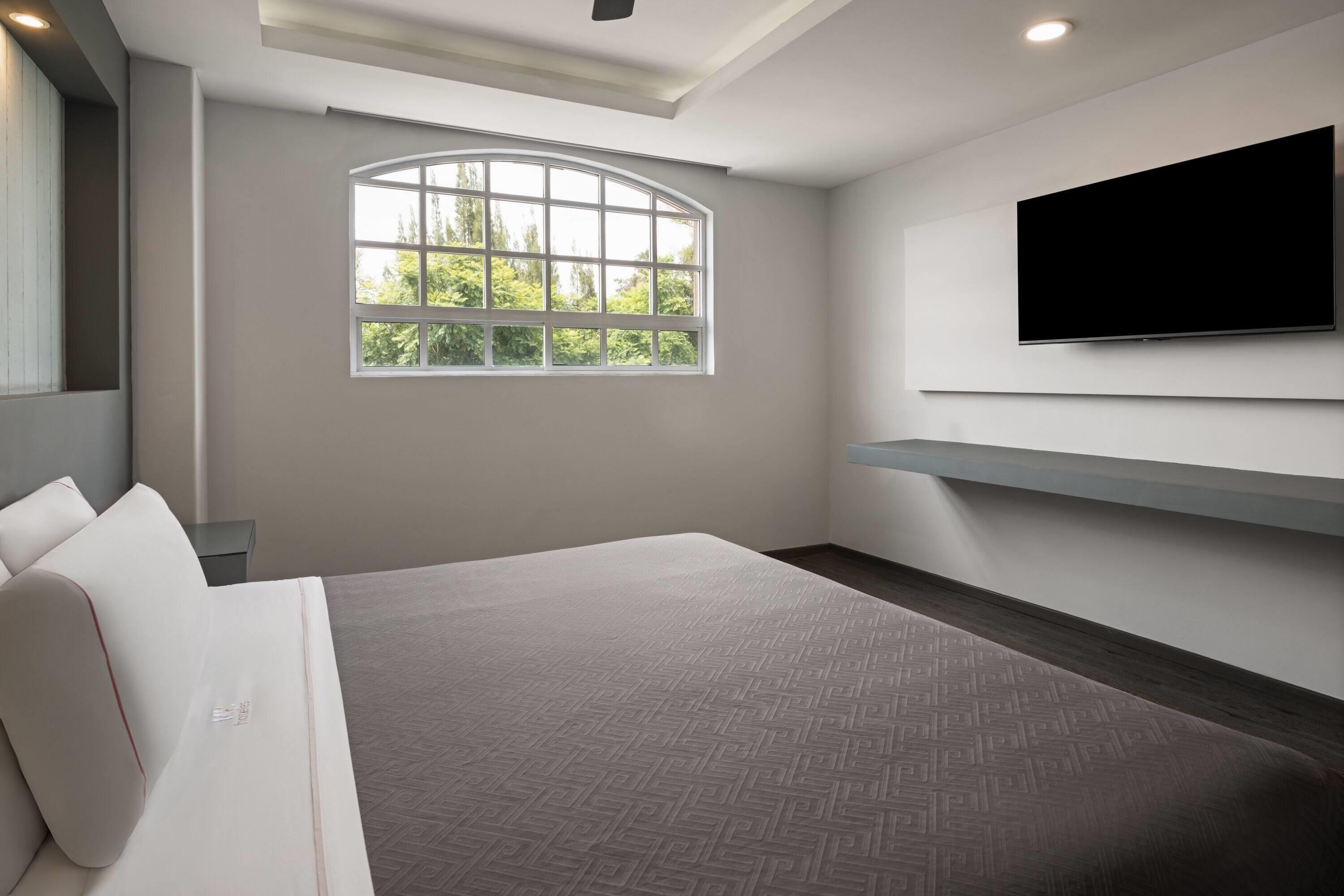 Quarto de hotel moderno com uma cama grande, televisão fixada na parede e janela com vista para o verde.