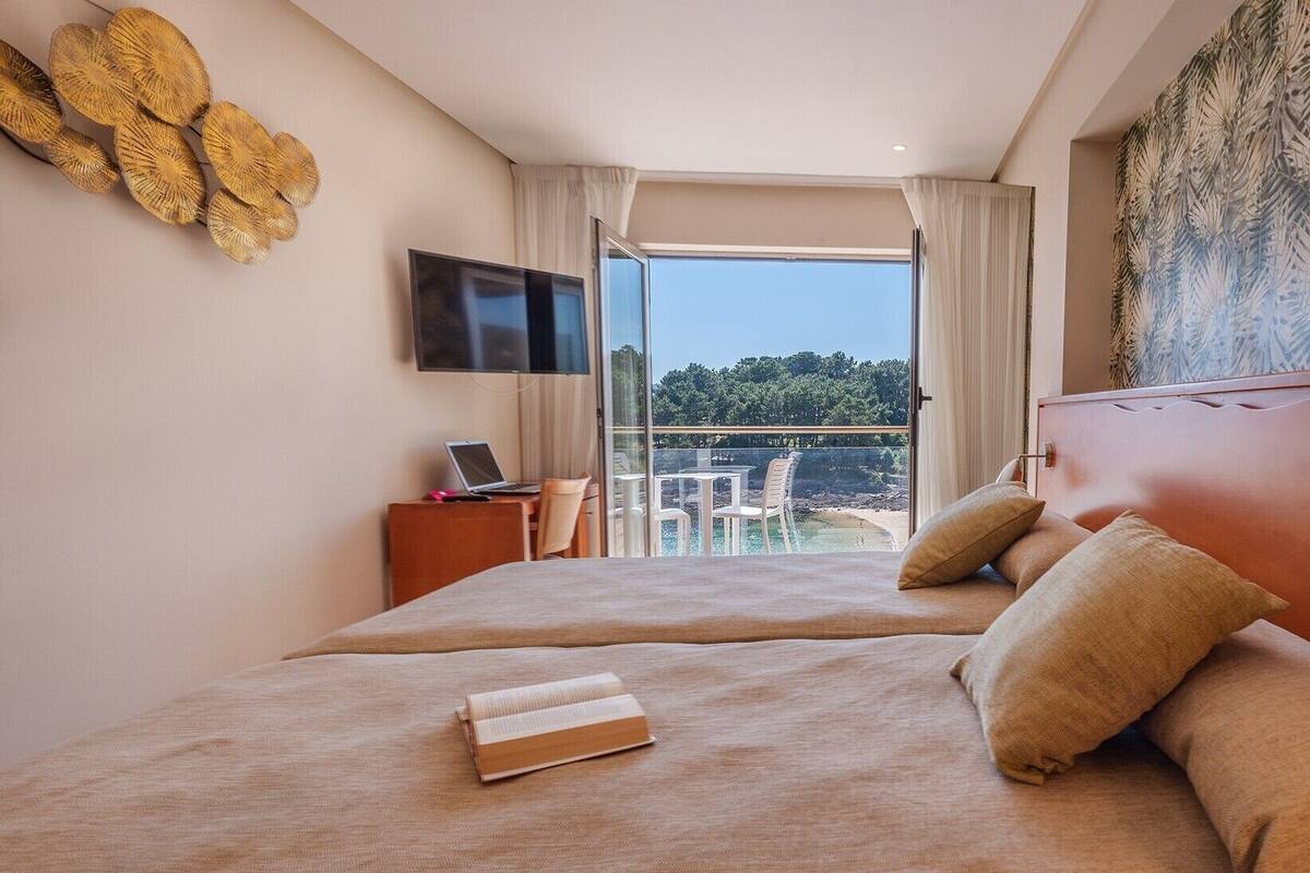 Quarto de hotel com duas camas, uma TV, um laptop sobre uma mesinha de apoio e uma varanda com vista para o mar.