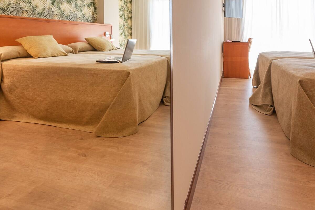 Quarto de hotel com duas camas, cada uma acompanhada de um laptop, piso de madeira e uma pequena mesa com abajur.