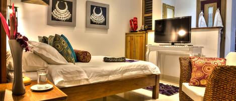 Quarto com cama, televisão, mesa de cabeceira de madeira, um vaso vermelho e duas molduras com colares na parede.
