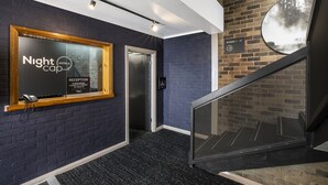 Uma área de recepção com a placa Night Cap, um balcão de atendimento, uma porta para os quartos 1 a 17 e uma escada com corrimão de metal.